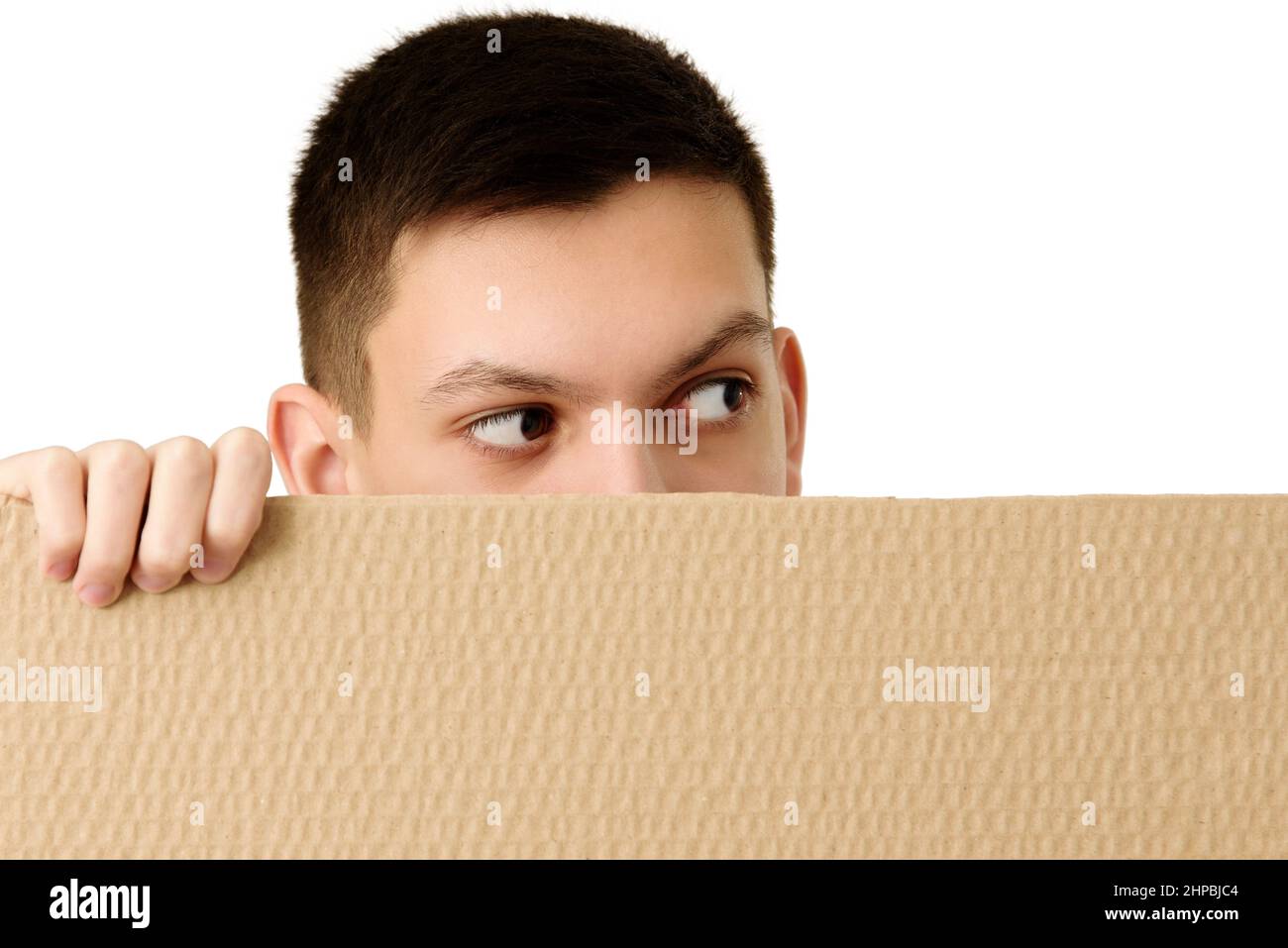 Teenage boy with sly eyes peeking out from behind blank brown cardboard ...