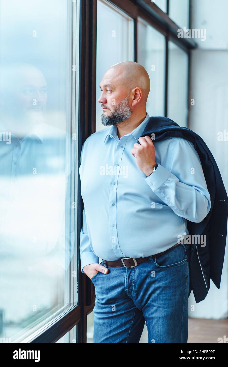 Portrait of mature bald man with beard staying near window looking away ...