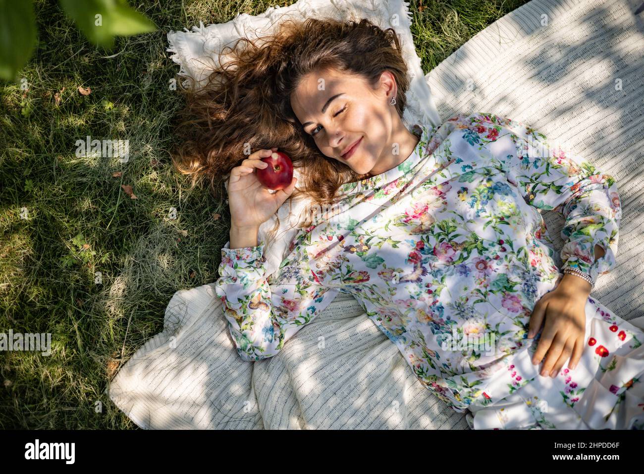 Portrait of beautiful woman posing lying on plaid at summer garden park. A book, fruit, a bag ...