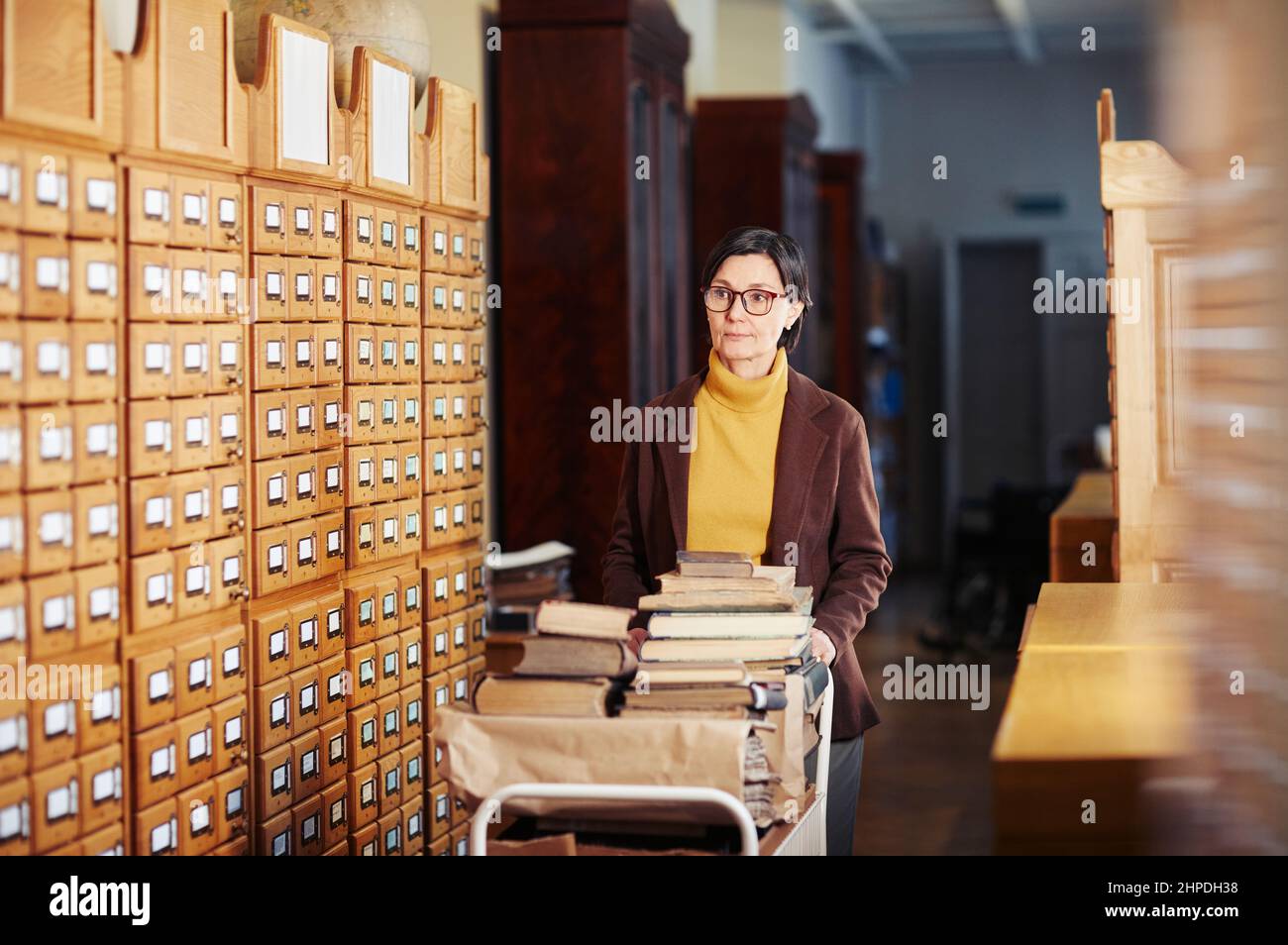 Warm toned portrait of female librarian pushing cart with books in ...