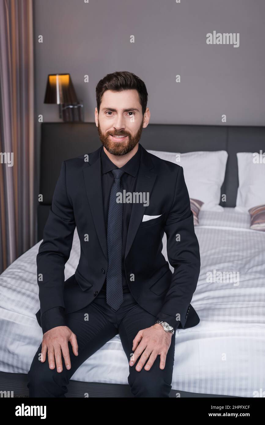 bearded man in suit smiling while sitting on bed in hotel room Stock ...