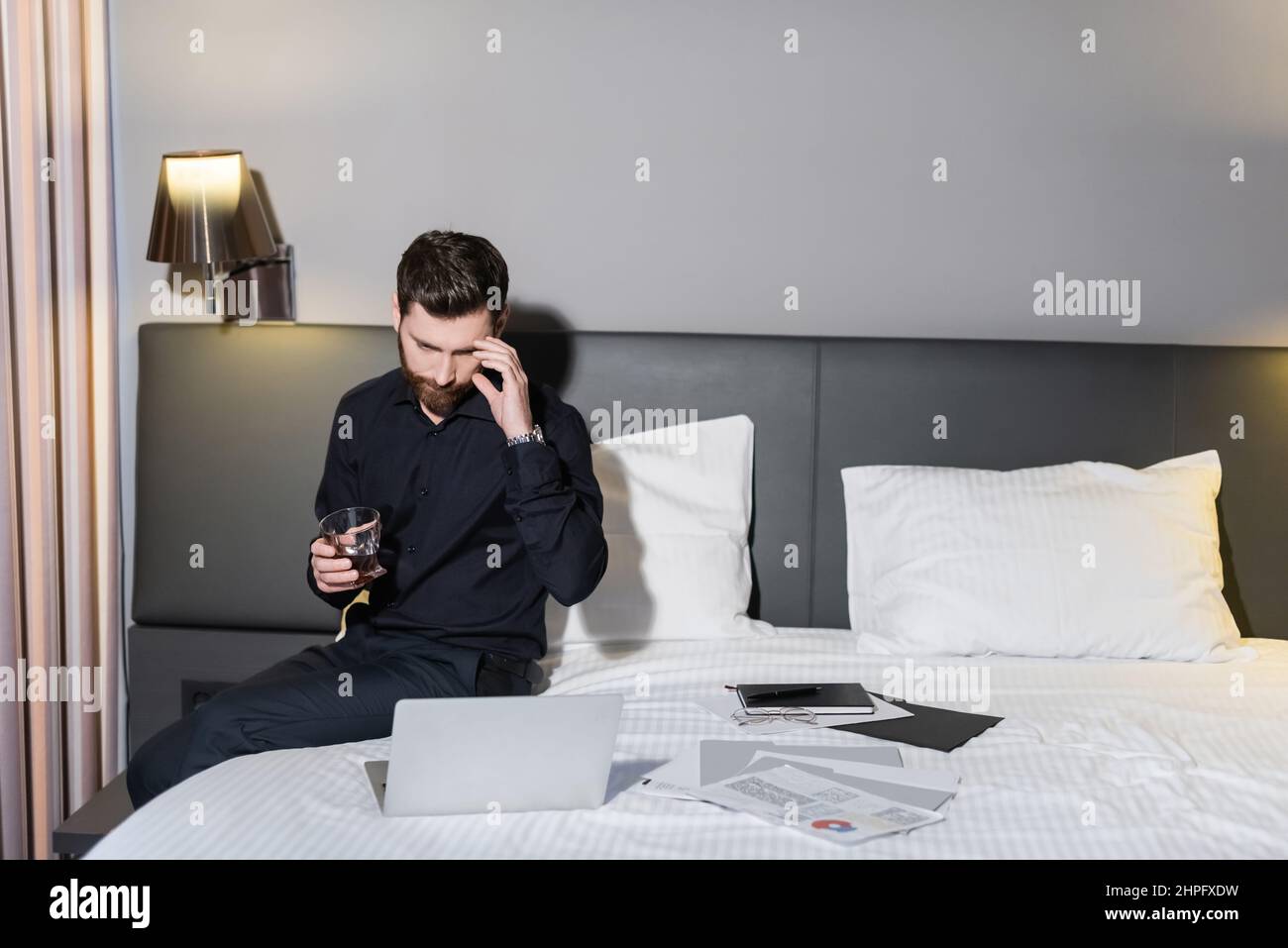bearded man holding glass of whiskey and looking at laptop in hotel ...