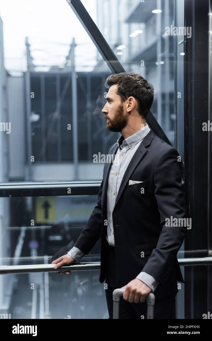 side view of bearded man in suit holding luggage handle in modern ...