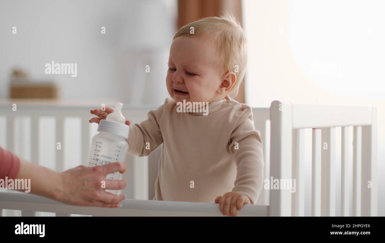 Cute little unhappy baby crying in crib, mother hand giving bottle with ...