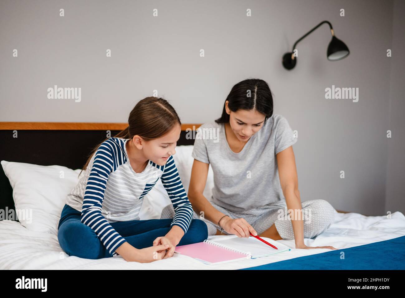 Nice beautiful mother and daughter doing homework while sitting on bed at home Stock Photo - Alamy