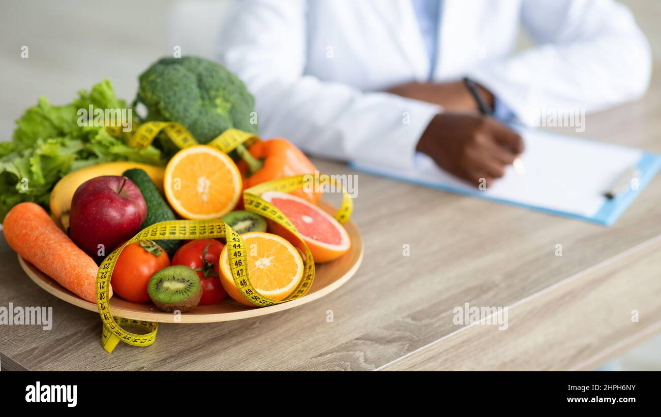 Black nutritionist writing diet plan, sitting at workplace in clinic ...
