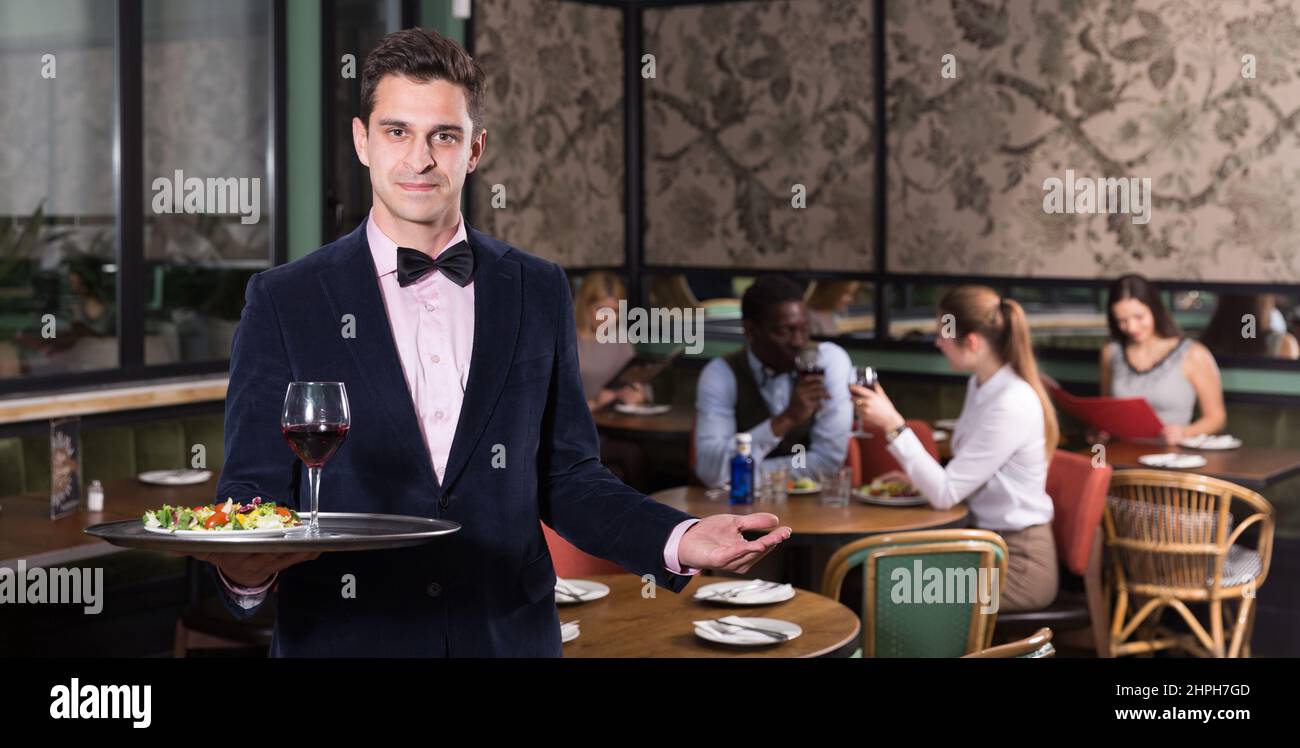Polite waiter holding tray at restaurant with customers behind him ...