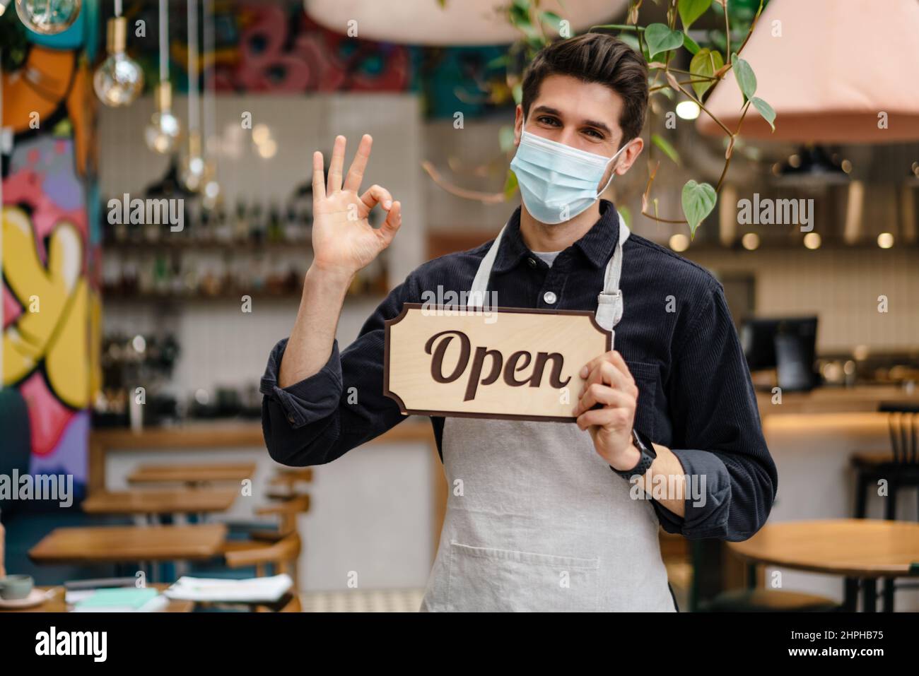 Young man waiter in apron showing open sign while standing indoors the ...