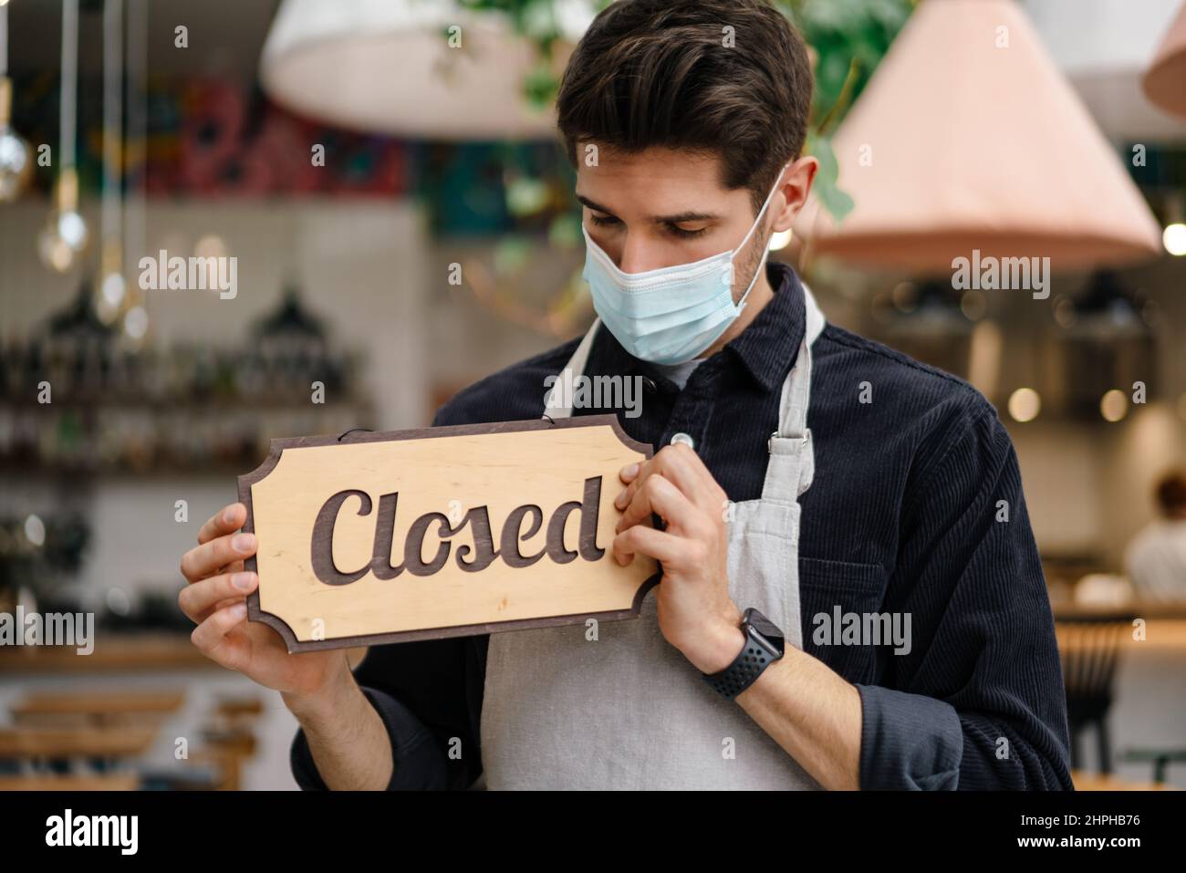 Young man waiter in apron and face mask showing closed sign while ...