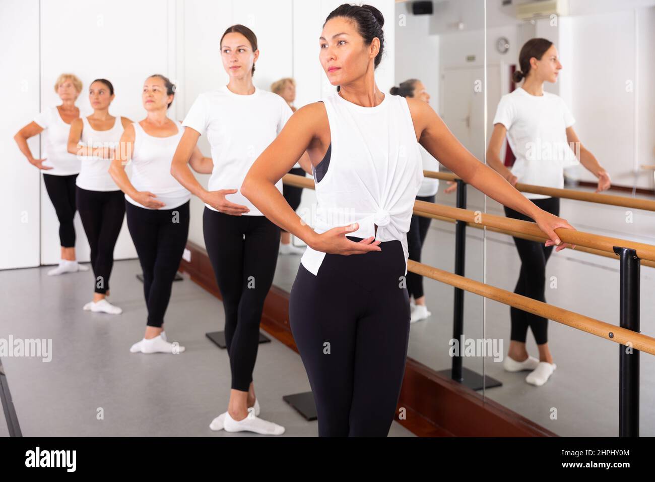 Group of women engaged in classical ballet stand holding onto a barre ...
