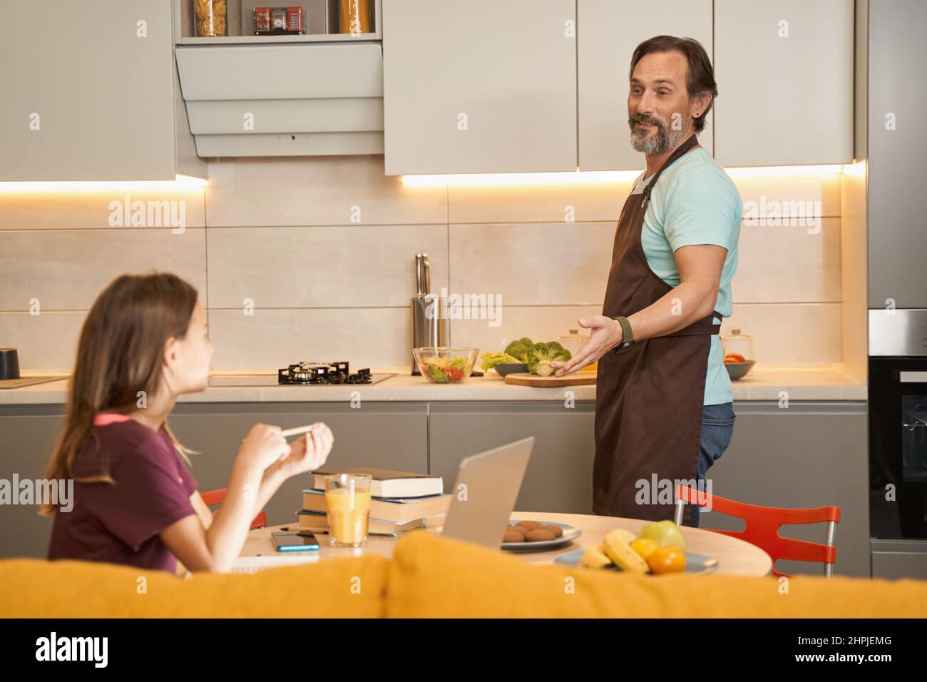 Adult man cooking in the kitchen while his daughter doing her homework ...