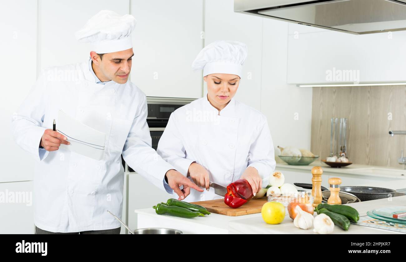 Female and male young cooks with paper recipe in uniform working Stock ...