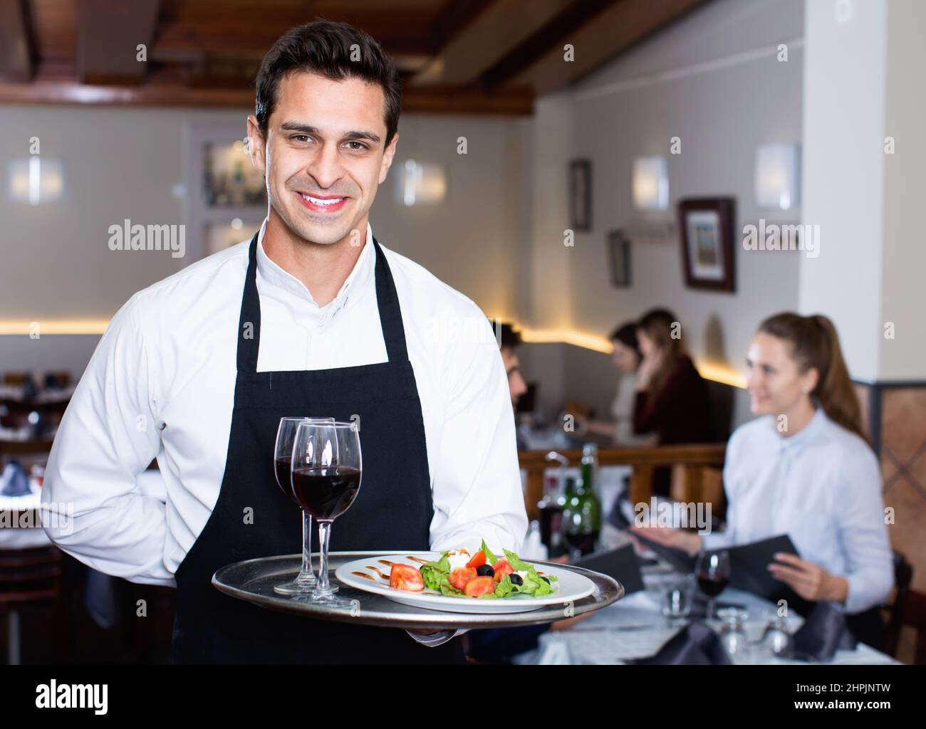 Portrait of smiling waiter with serving tray meeting restaurant guests ...