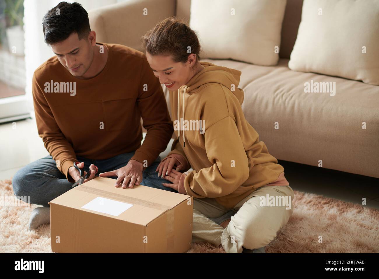 Young man opening the parcel with scissors with excited woman sitting ...