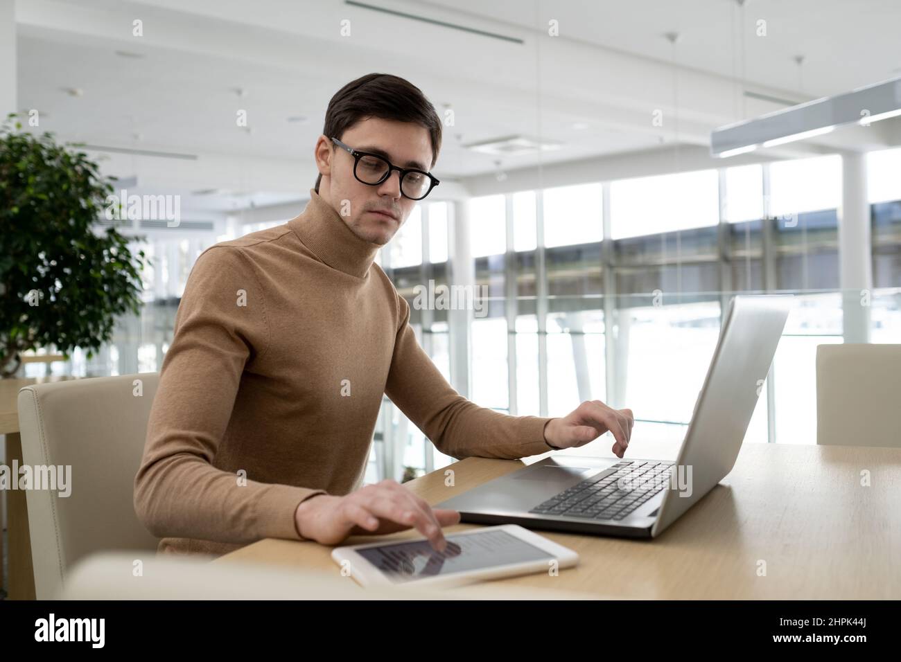 Young serious high-tech developer using tablet and laptop while sitting ...