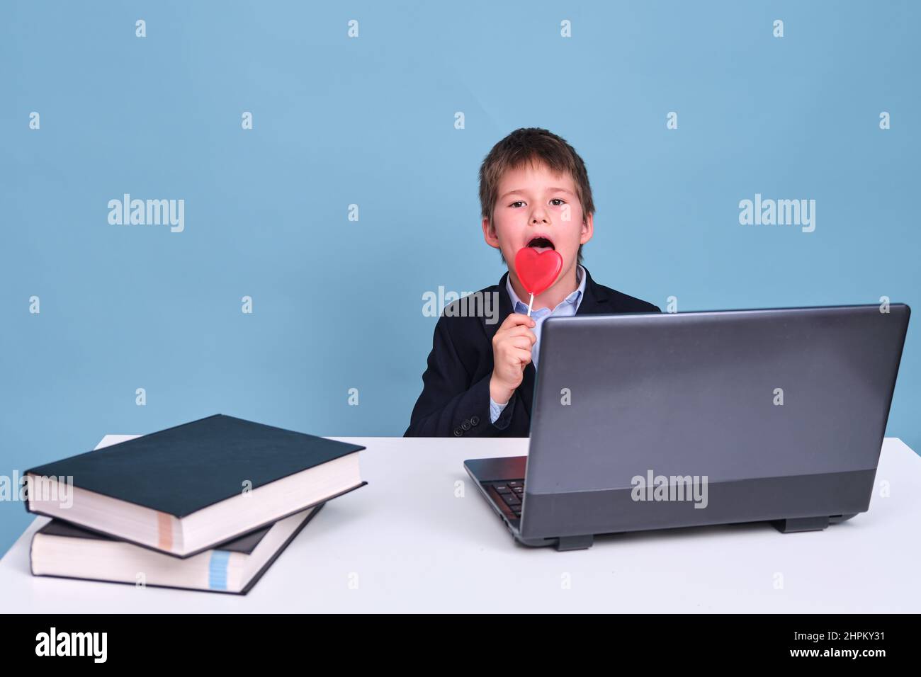 A boy in a school suit eats a red candy on a stick at a computer during ...