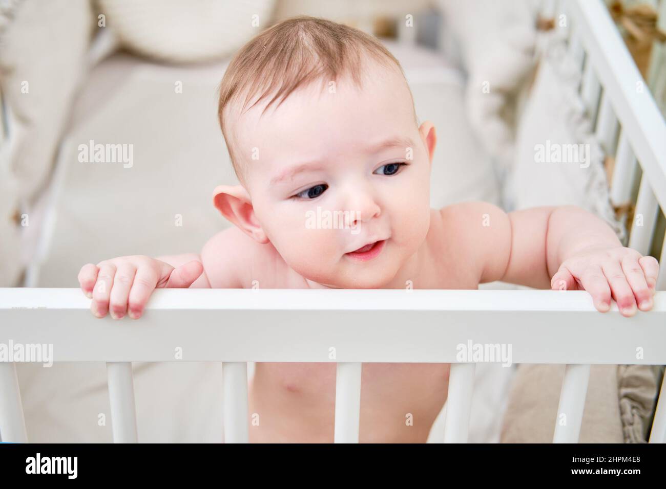 A curious baby is standing in a crib, holding the handrails and smiling ...