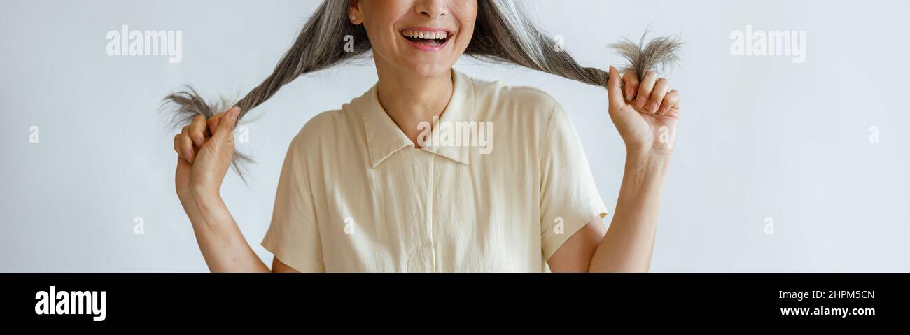 Happy middle aged Asian woman holds twisted hoary hair on light grey ...