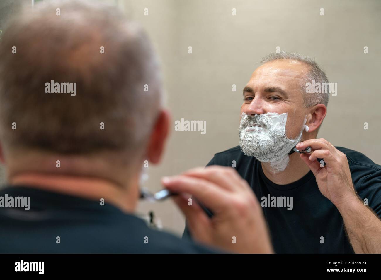 Handsome white-haired beared man shaving off his beard looking in a ...