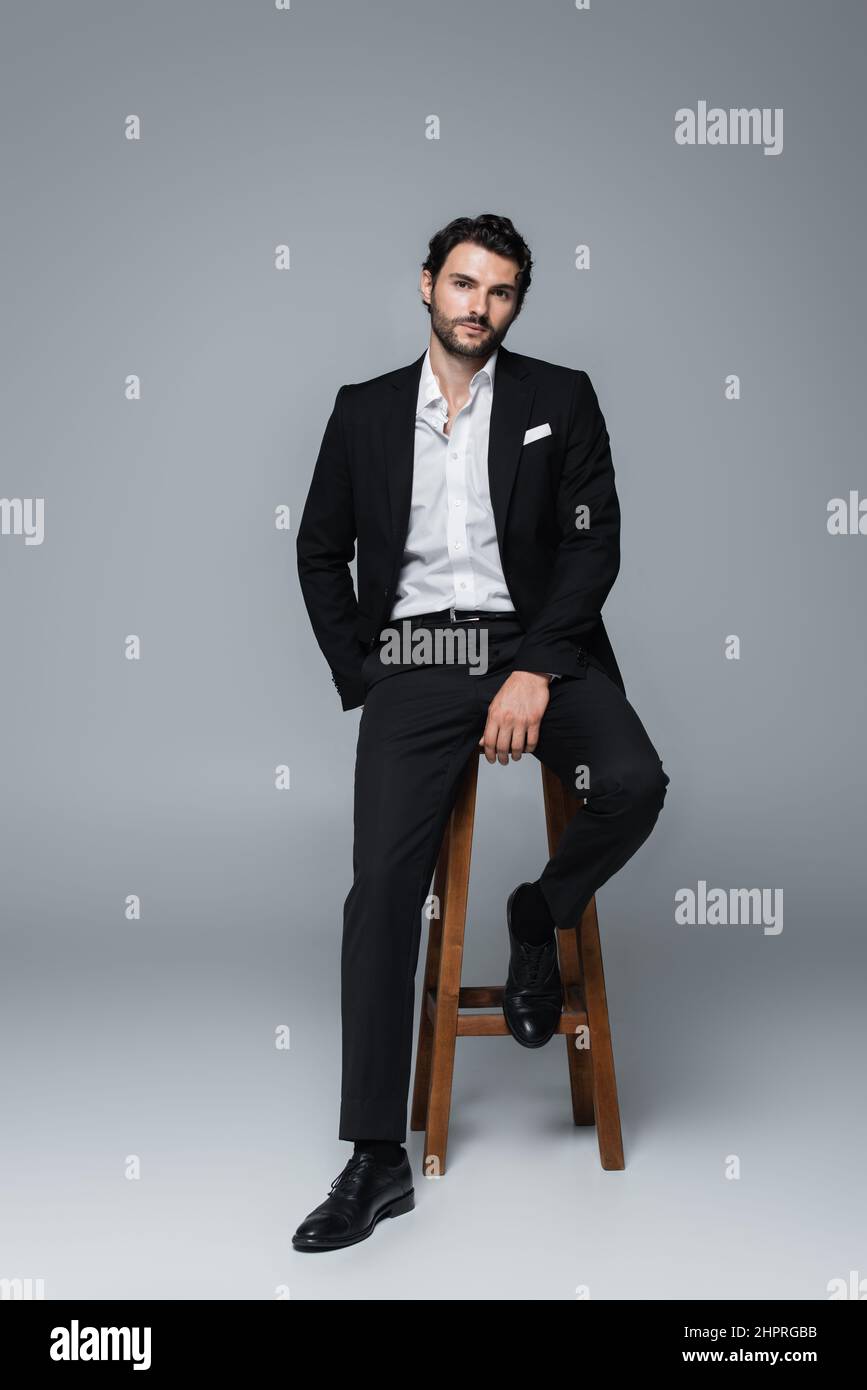full length view of man in black suit sitting on high stool and looking ...
