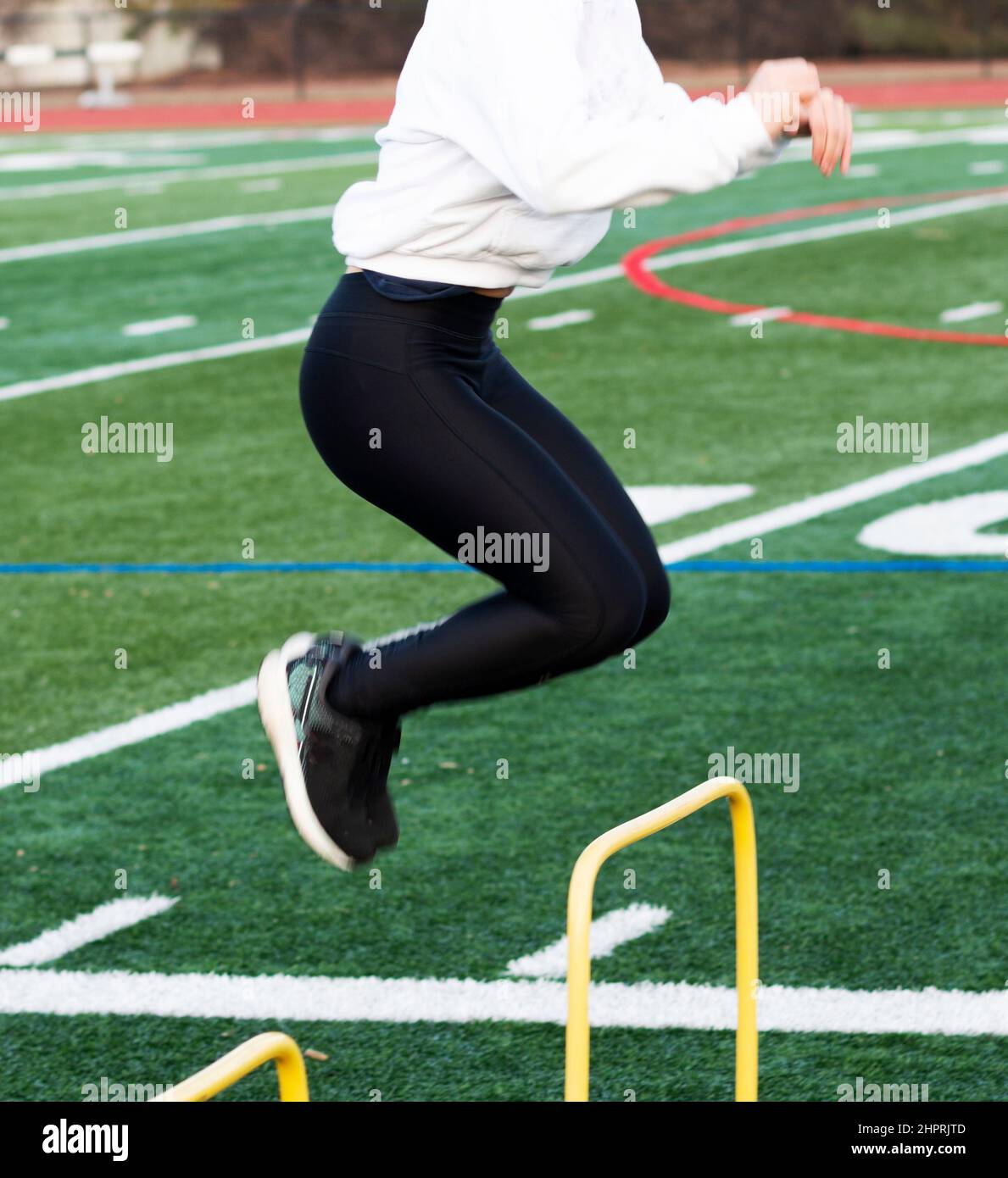 A high school girl is jumping over plastic yellow hurdles on a turf ...