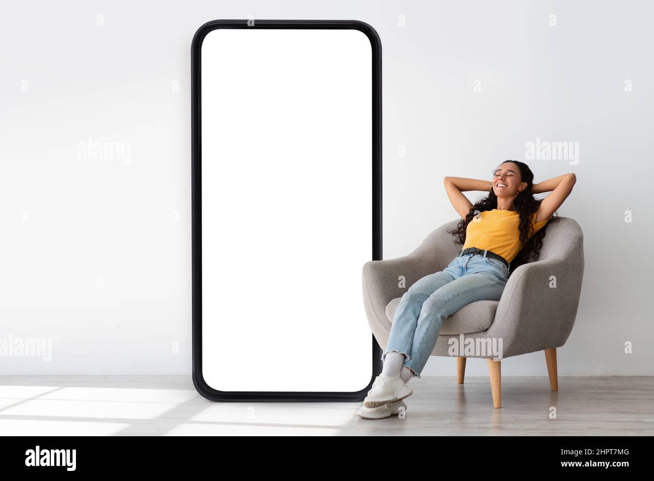 Happy young woman relaxing in armchair nar big smartphone with blank ...