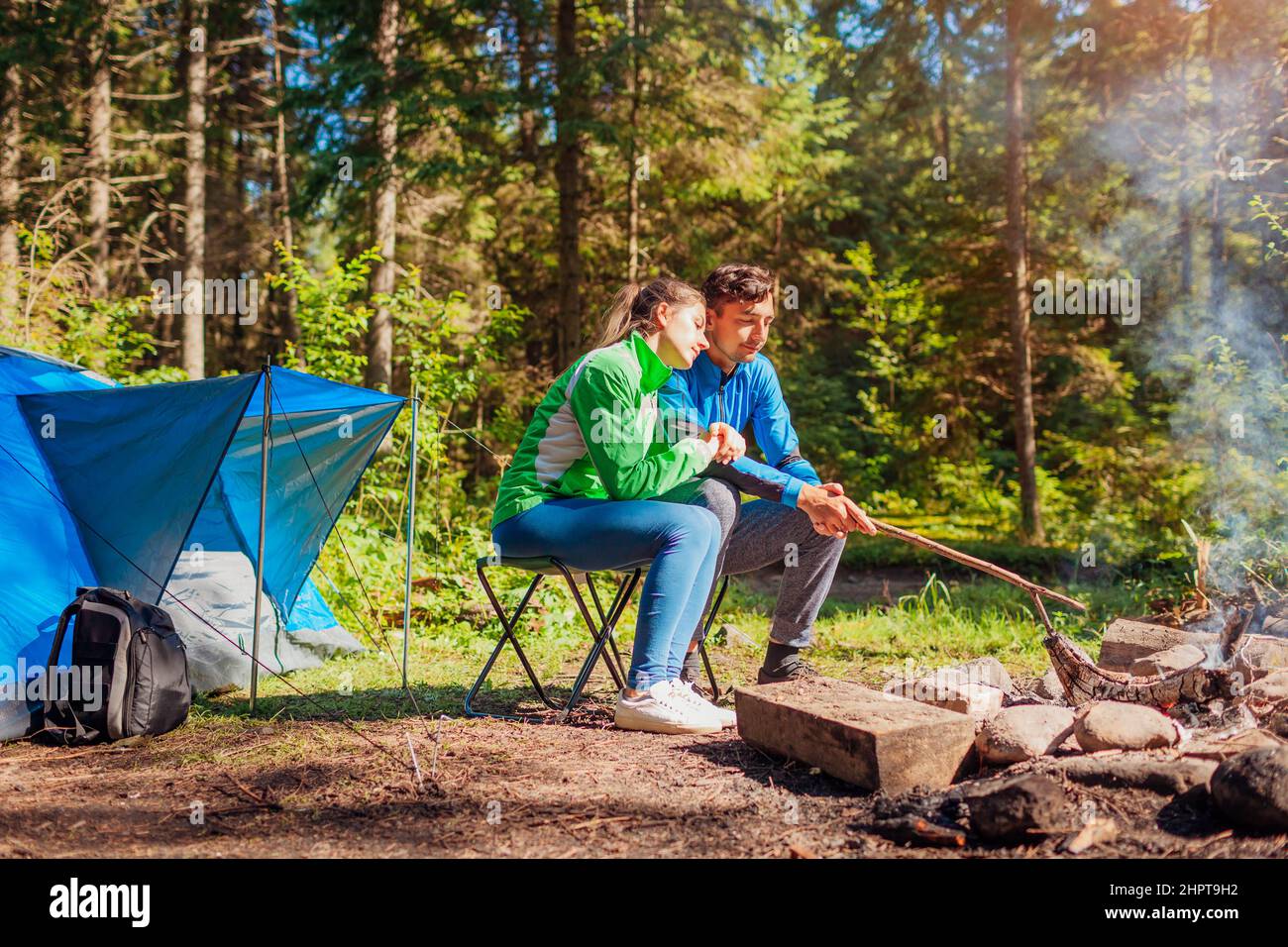 Young couple of travelers enjoy campfire in summer forest. Tourists ...