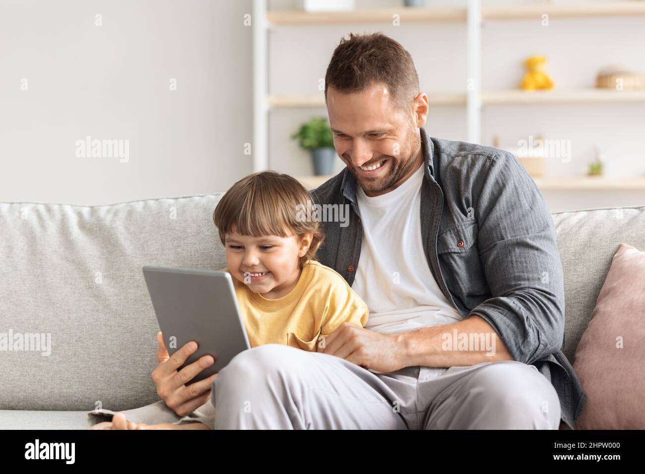 Young father and little son video chatting via digital tablet, sitting on sofa and smiling to ...