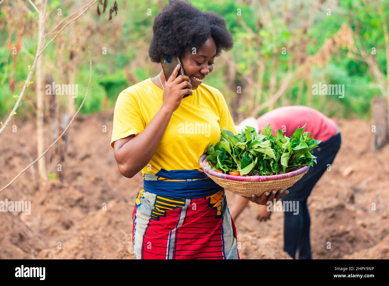 Female African farmer making phone call while carrying basket of ...