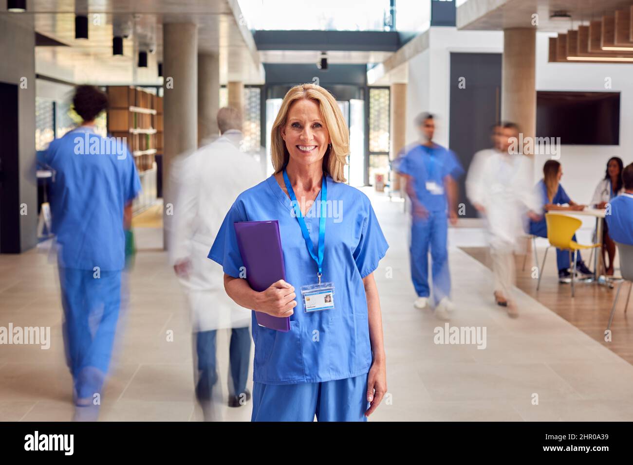 Portrait Of Female Doctor Wearing Scrubs Holding Patient Notes In Busy ...