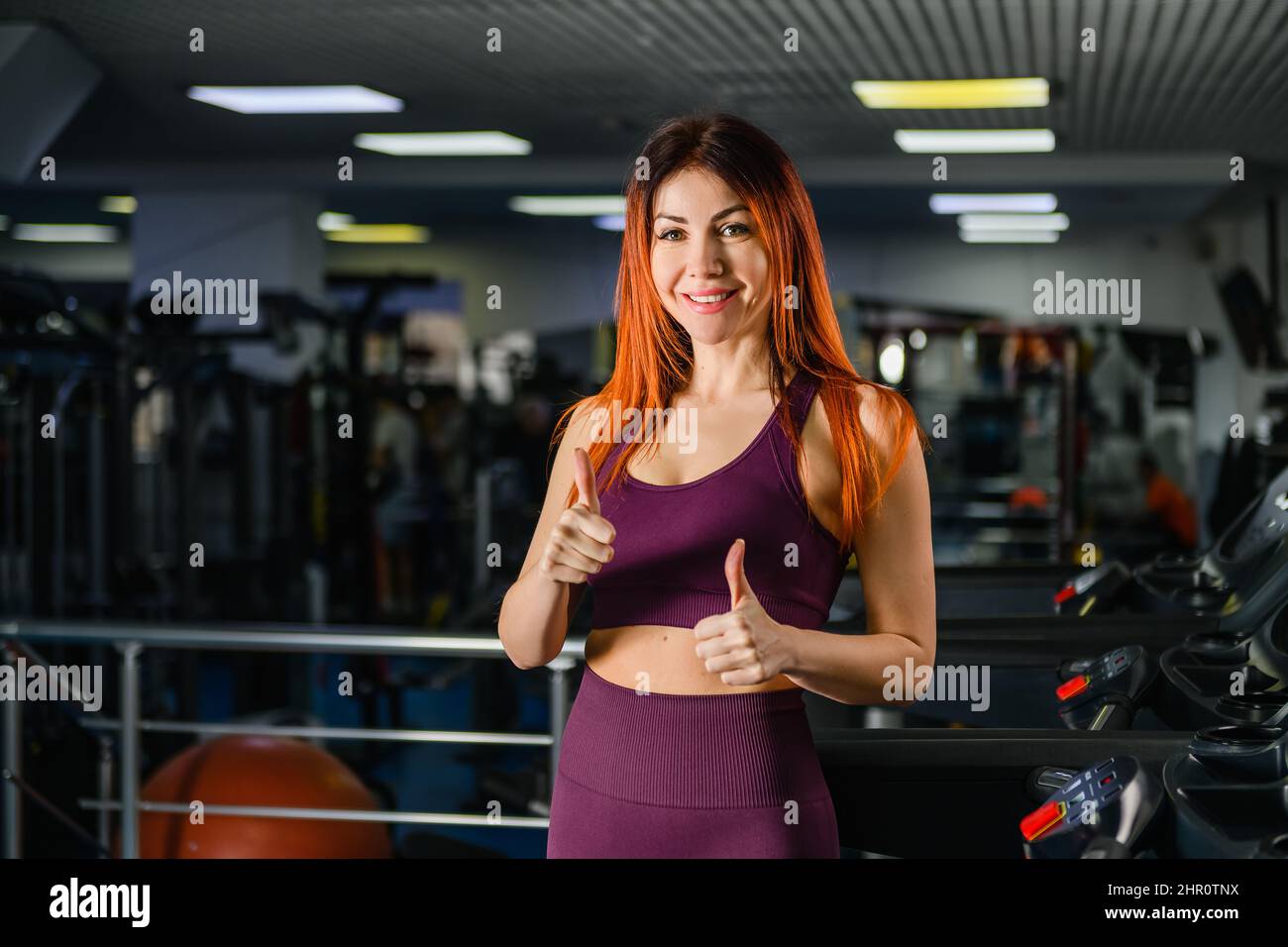 Sportive fit woman show thumb up gesture in gym interior near treadmill ...