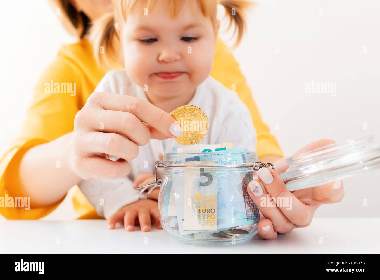 Baby girl and her mother puts bitcoin in a glass jar with currency ...