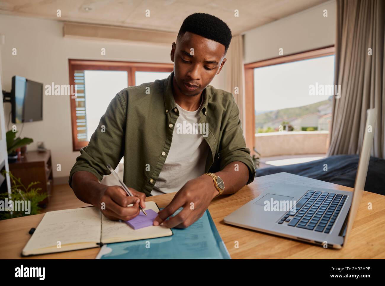 Young adult black male writing note on sticky note next to laptop ...