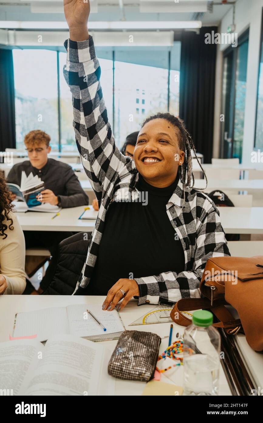 Cheerful female student raising hand while sitting at desk in classroom ...