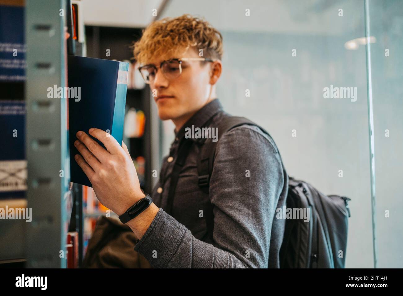 Side view of young man wearing eyeglasses searching book in library at ...