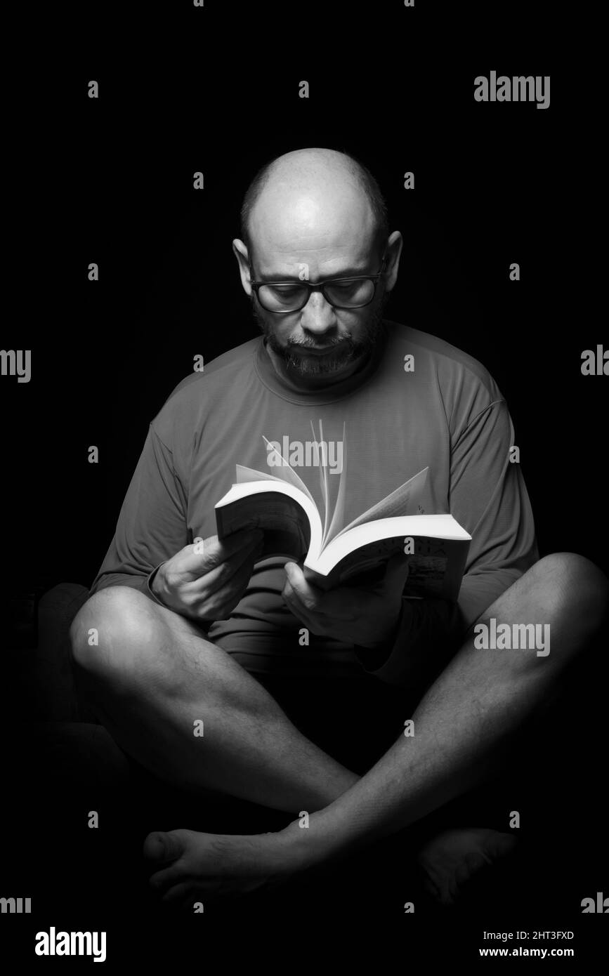 Portrait of a bearded, bald man reading a book against black background ...