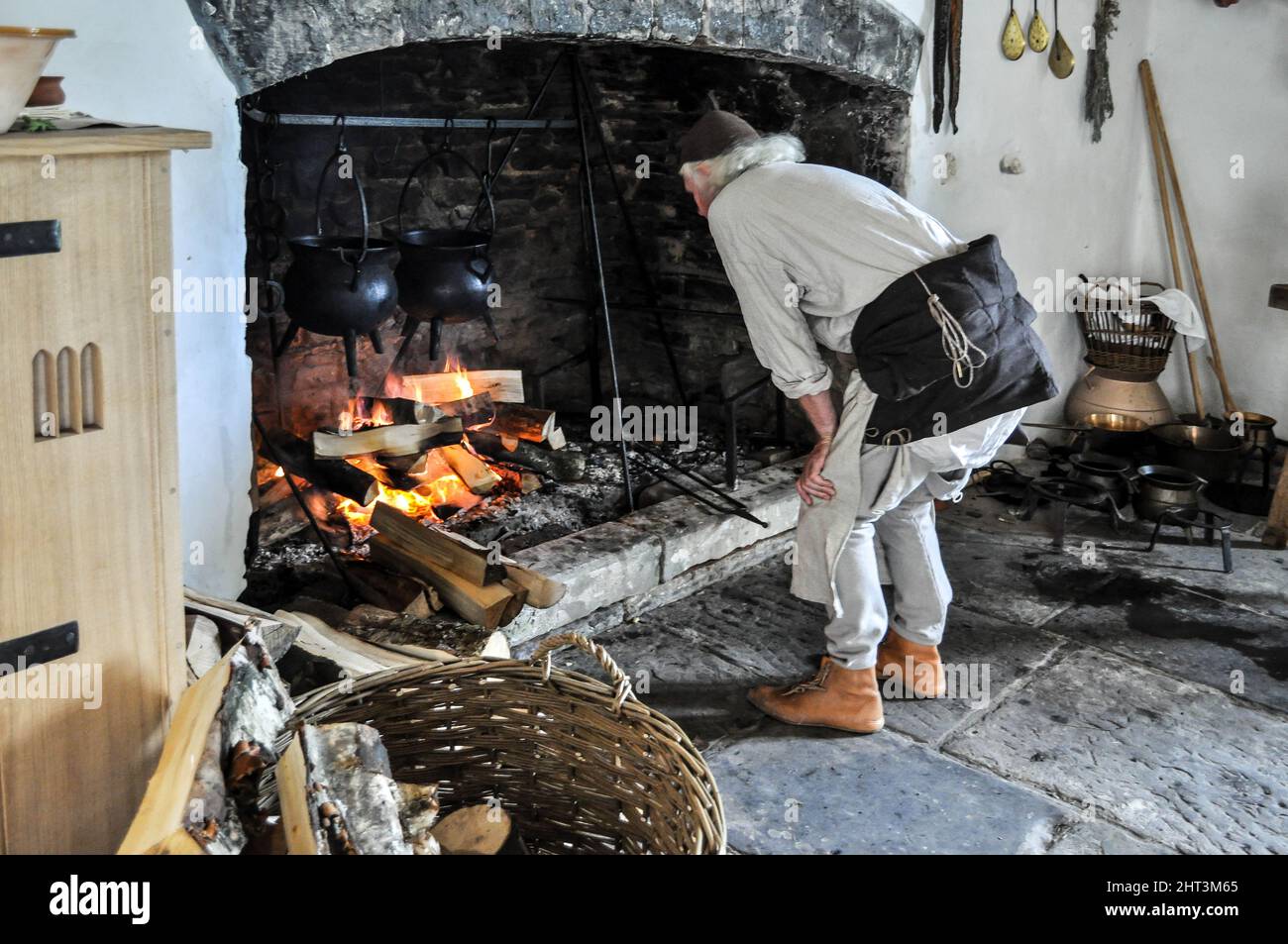 Old man leaning over a medieval oven with two cauldrons hanging in it ...