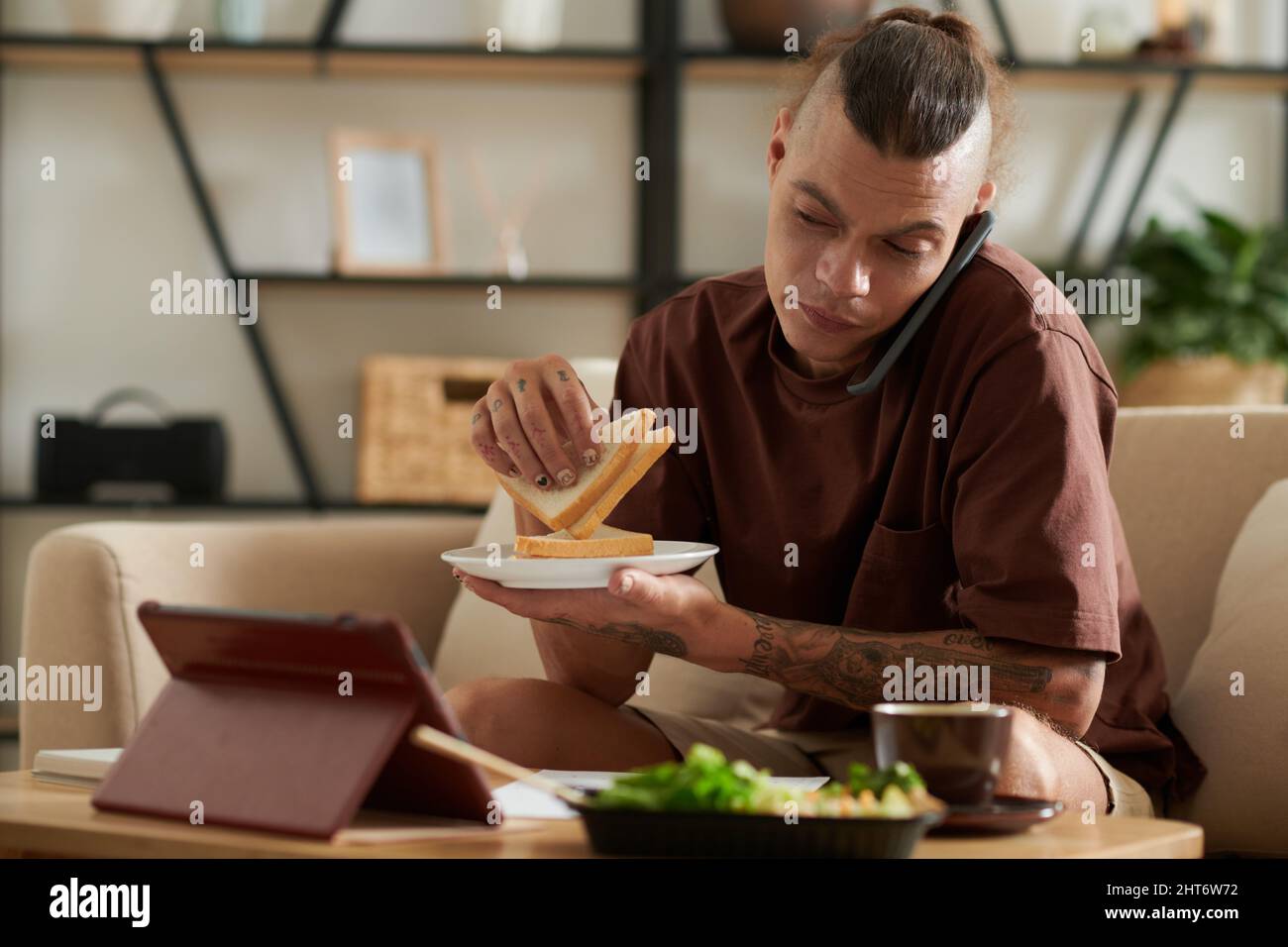 Serious young man eating sandwich and talking on phone with colleague ...