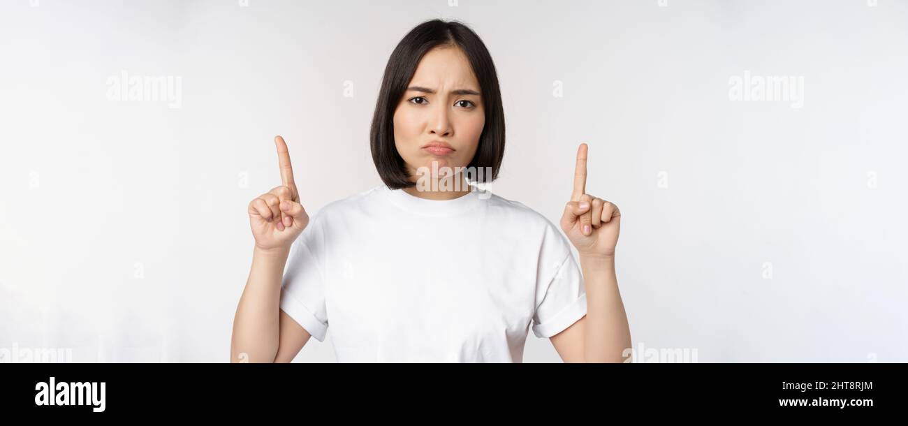 Sad and disappointed young asian woman pointing fingers up, showing ...