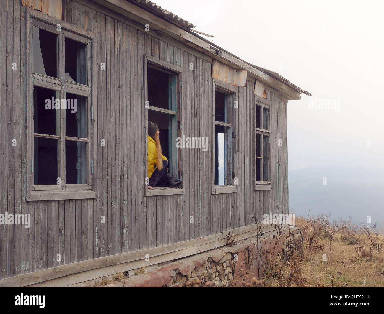 A girl sits in the window opening of an old wooden abandoned house with ...