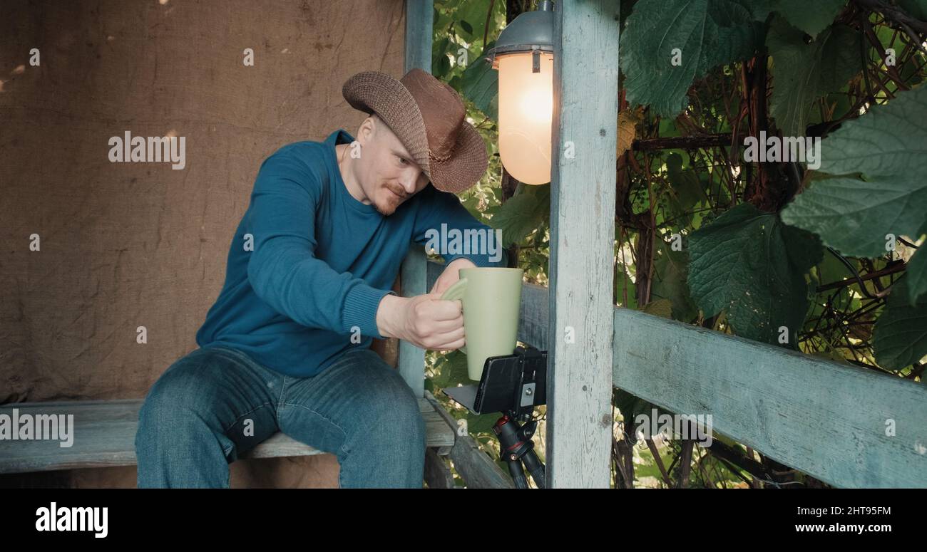 Cowboy man on porch drinks coffee from a mug and communicates online by ...
