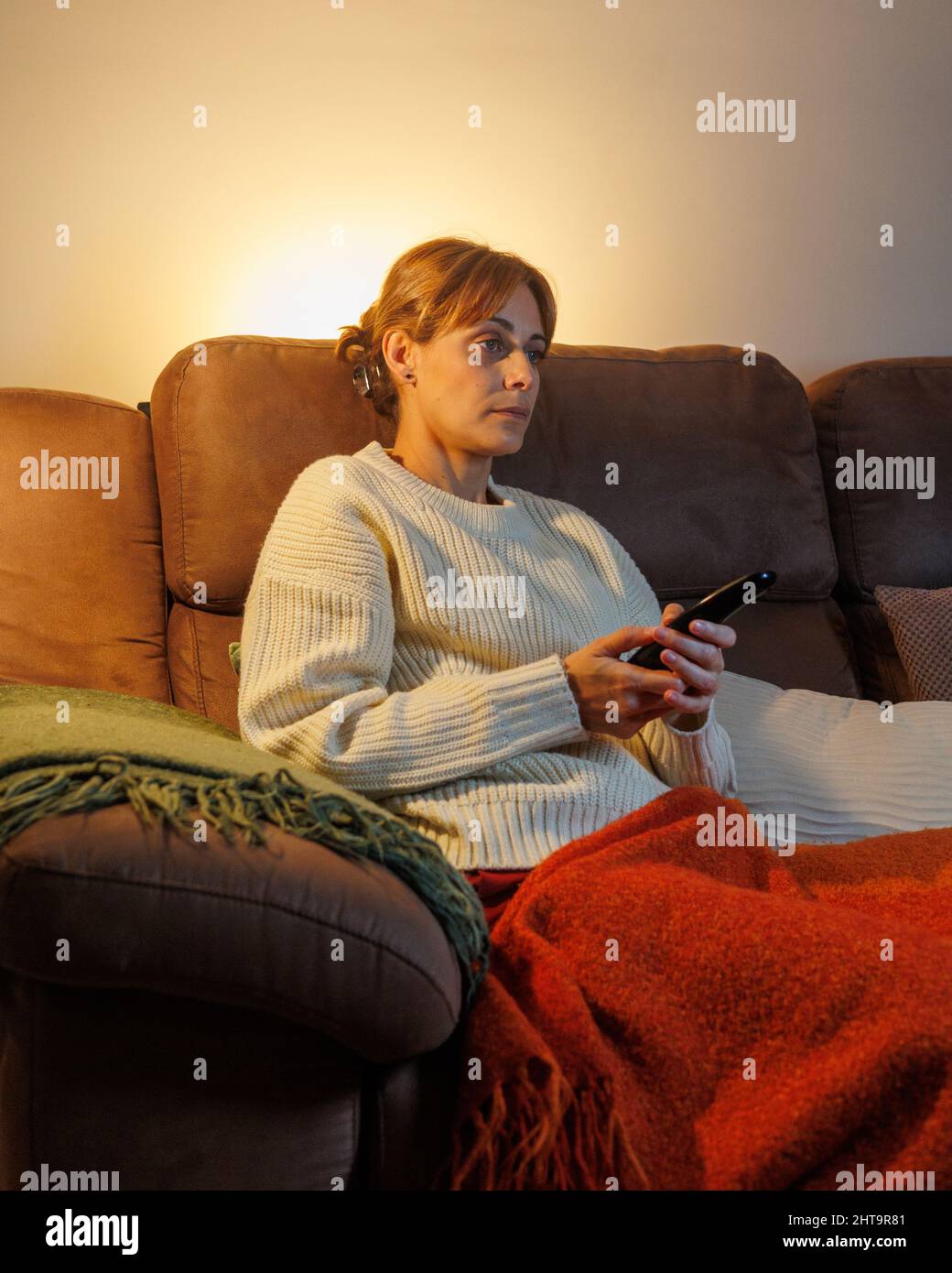 Woman in white sweater sitting on sofa and holding remote control in ...