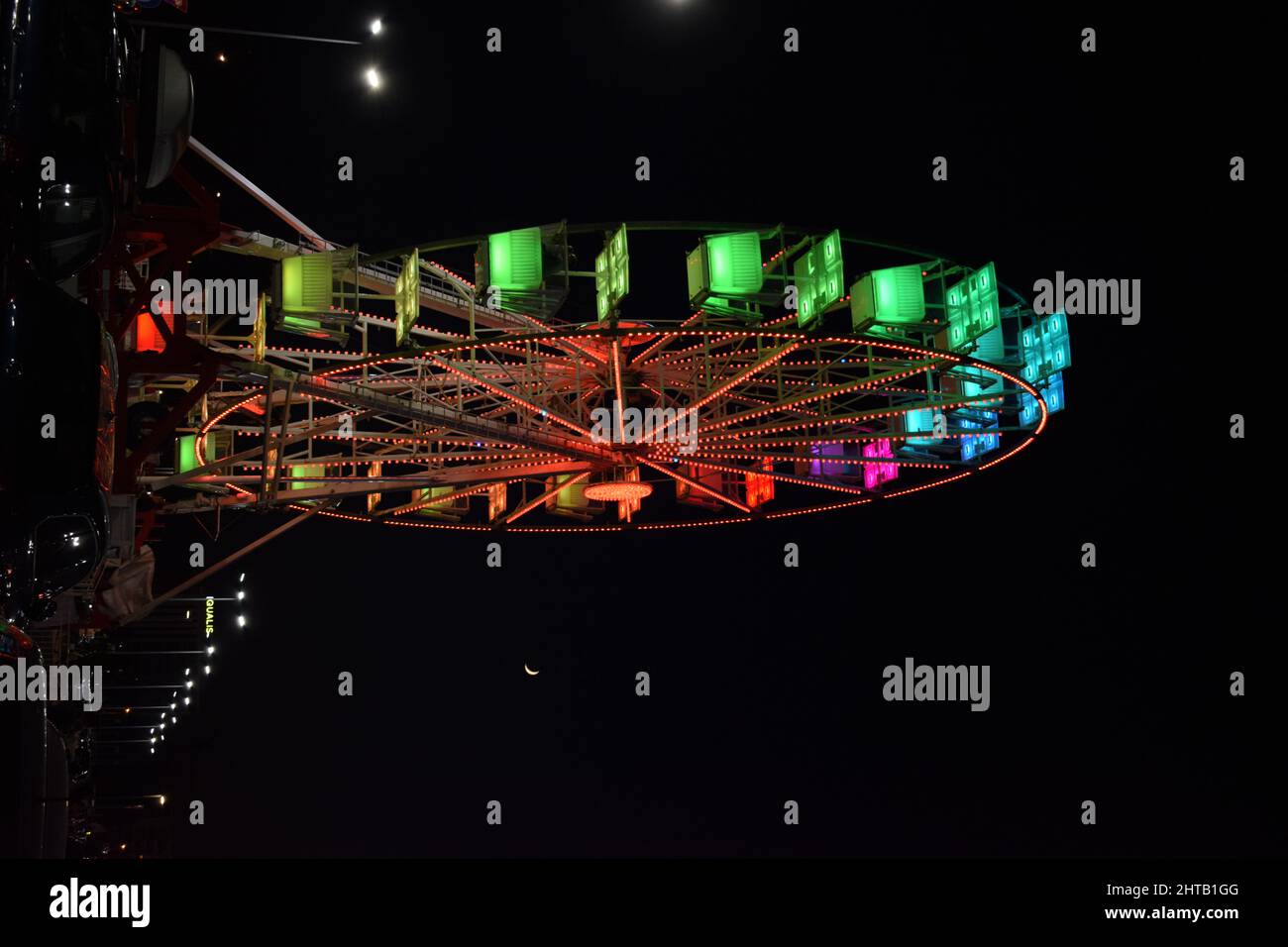Vertical night view of a colorful ferris wheel, and cars beneath it ...