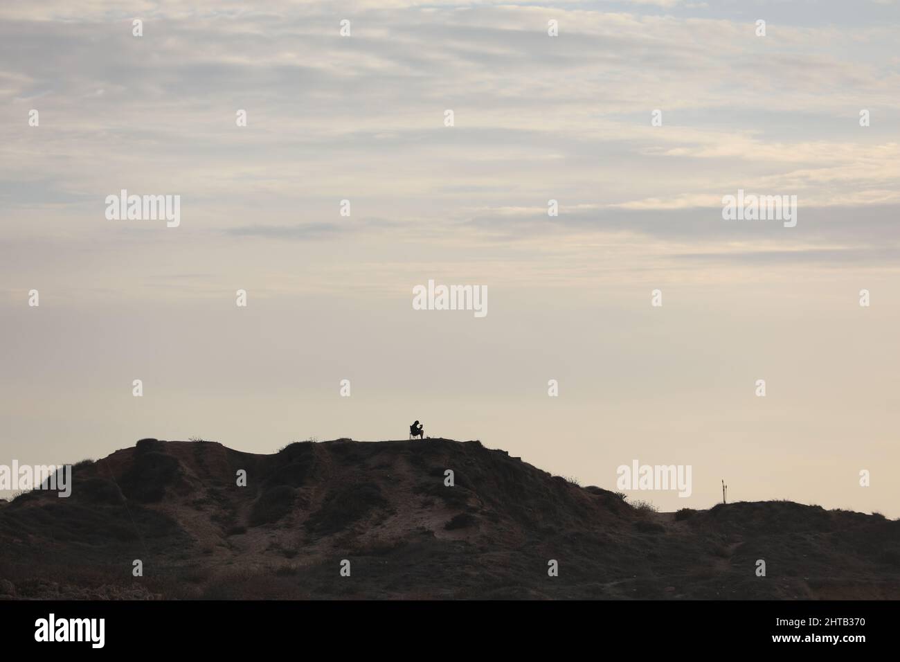 silhouette of a man in a cap sitting on a mountain watching the sunset ...