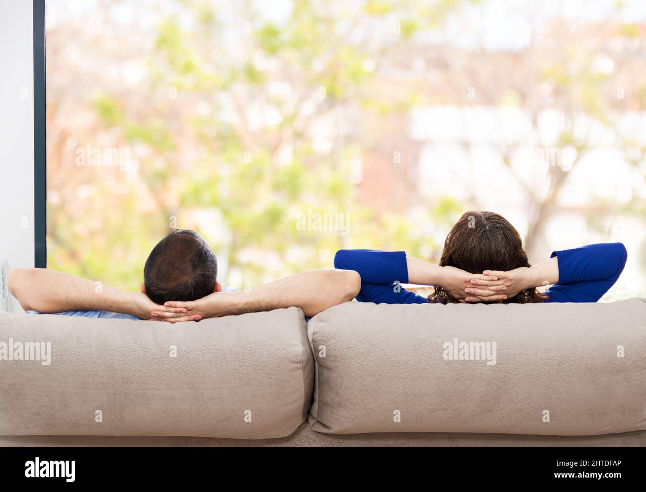 Rear view of a couple relaxing on a sofa at home and looking outside a ...