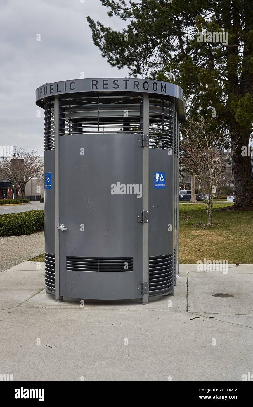 A round grey outdoor public restroom on concrete in front of a tree and ...