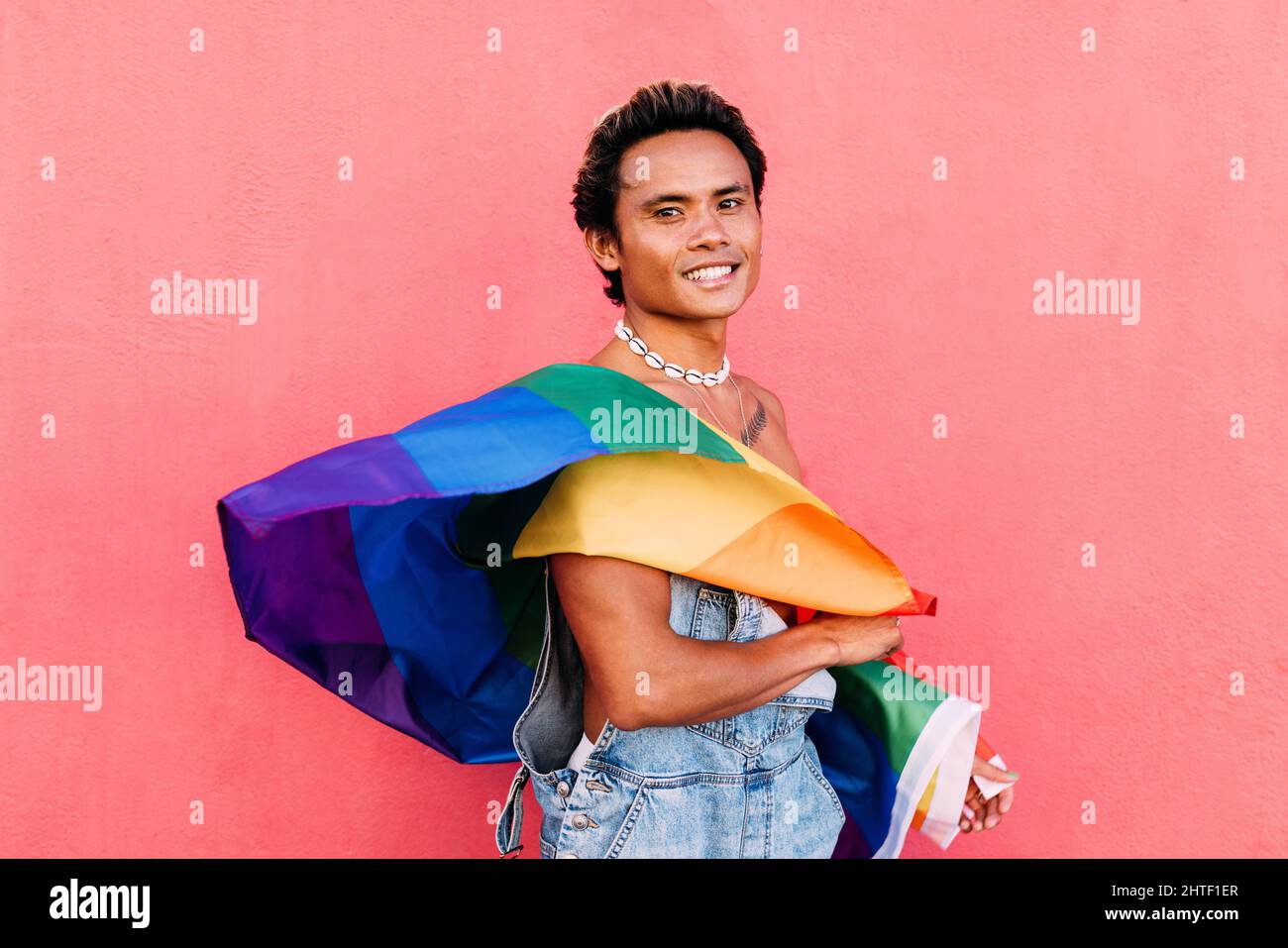 Portrait of a young smiling guy posing with LGBT flag against pink wall ...