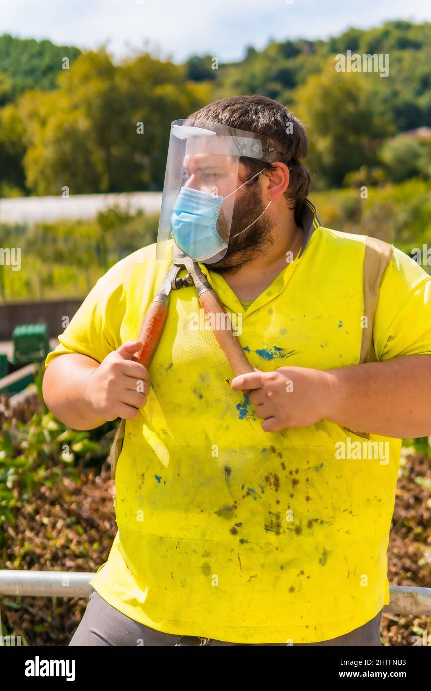 A gardener with a bush clipper wearing a protective face shield and ...