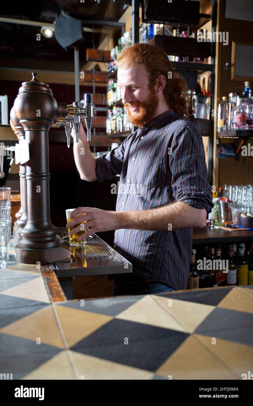 Side view of optimistic male barkeeper with red hair pouring beer on ...