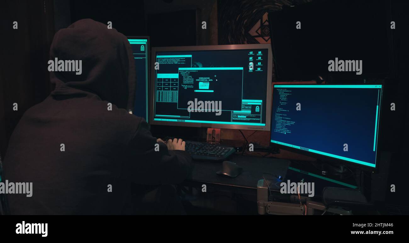 Hacker in a hood sits in front of computer screens and hacks databases ...